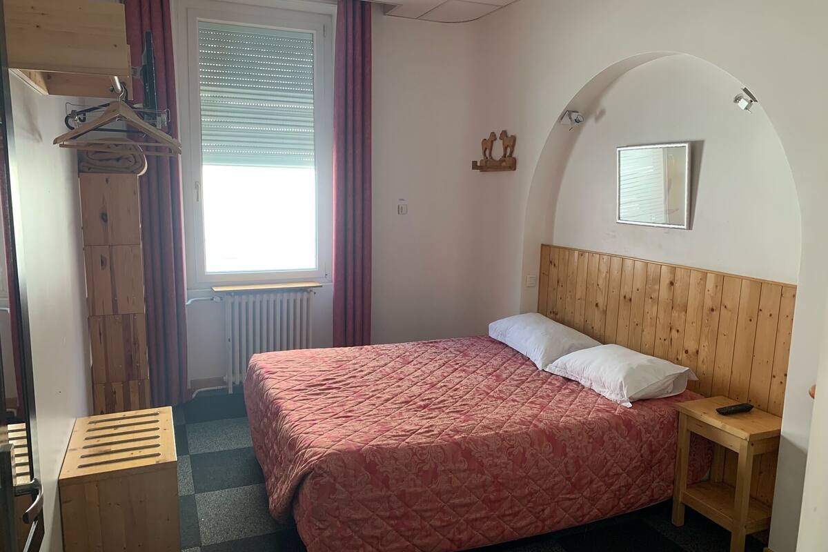 Quarto com cabeceira de madeira, cama com colcha vermelha, mesa de cabeceira de madeira com controle remoto, guarda-roupa de madeira, radiador e janela com persianas.