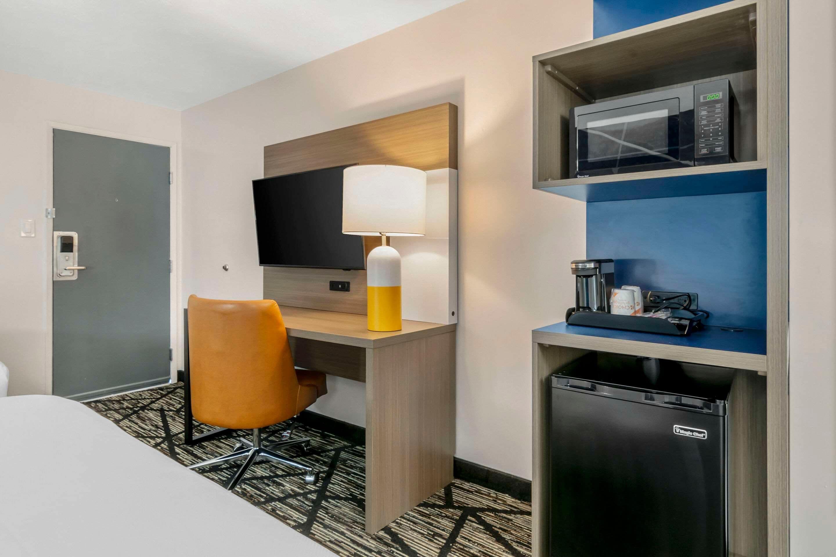 Quarto de hotel com cama, escrivaninha com luminária, televisão, micro-ondas, máquina de café e mini-geladeira.