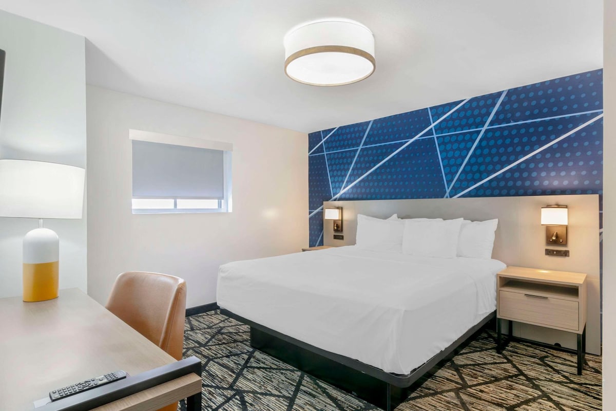 Quarto de hotel com cama bem arrumada, uma escrivaninha com controle remoto, uma luminária e um tapete estampado.
