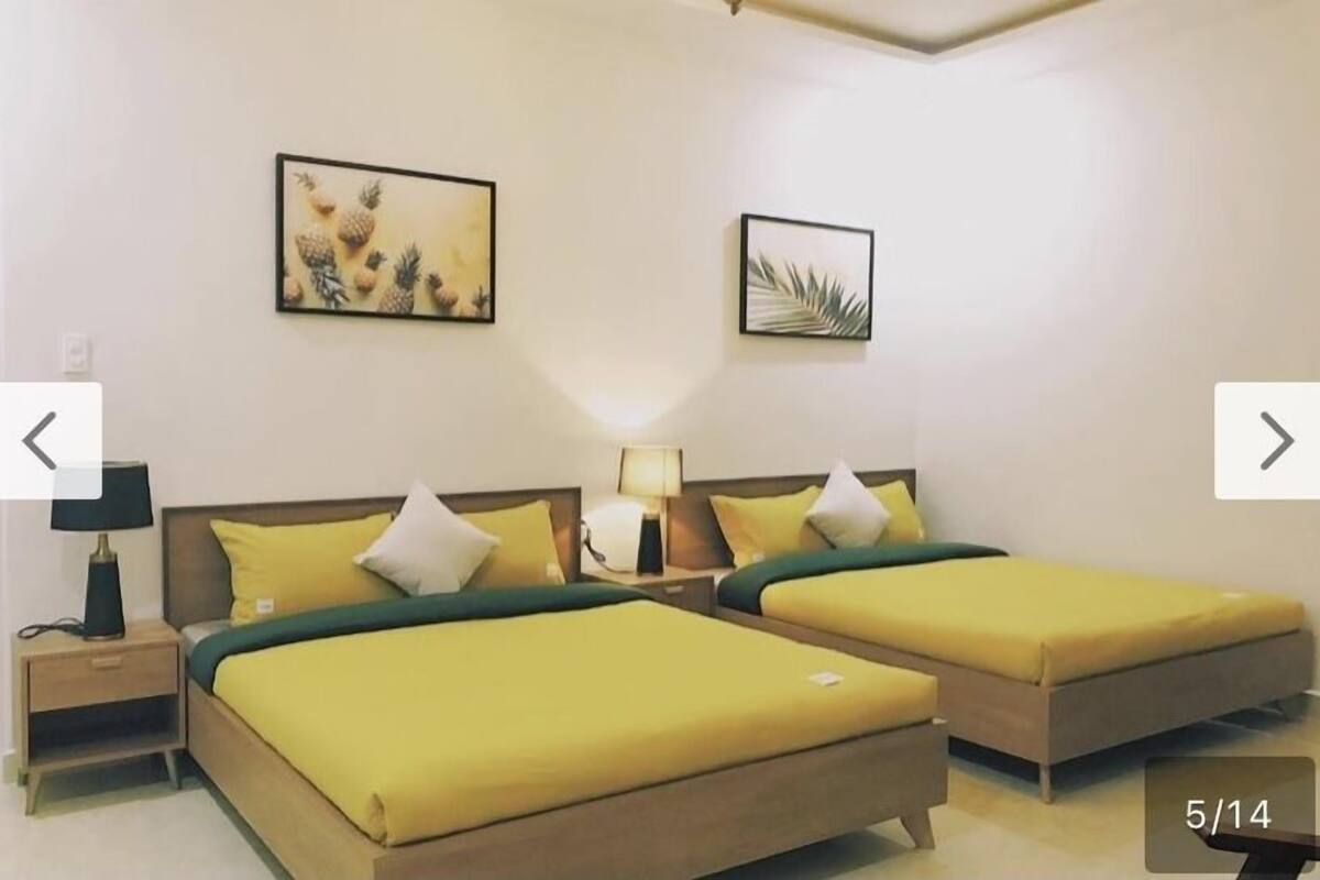 Quarto de hotel com duas camas, cada uma com roupa de cama amarela e almofadas verdes. Há duas mesas de cabeceira com luminárias, e as paredes são decoradas com obras de arte emolduradas.