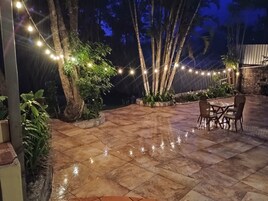 Um pátio externo com piso de azulejos, uma mesa com cadeiras, luzes de festa e plantas em vasos.