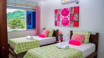 Quarto de hotel com duas camas, cada uma com colcha florida e almofadas cor-de-rosa. Há uma janela com vista para áreas verdes.