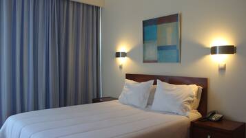 Quarto de hotel com cama, duas luminárias de cabeceira, mesa de apoio com telefone e um quadro na parede.