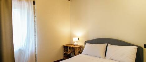 Uma cama bem arrumada com roupa de cama e almofadas brancas, uma mesa de cabeceira de madeira com abajur e uma janela com cortinas.