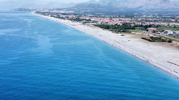 Vista aérea de uma área costeira com uma extensa praia de areia, um horizonte urbano ao fundo e montanhas à distância.