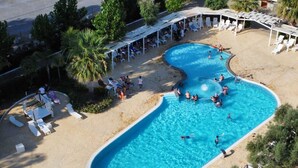Uma grande piscina ao ar livre rodeada por espreguiçadeiras e guarda-sóis, com uma área de estar coberta e palmeiras.