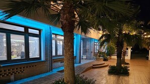 Área externa bem iluminada com um caminho pavimentado, palmeiras e um edifício com grandes janelas.