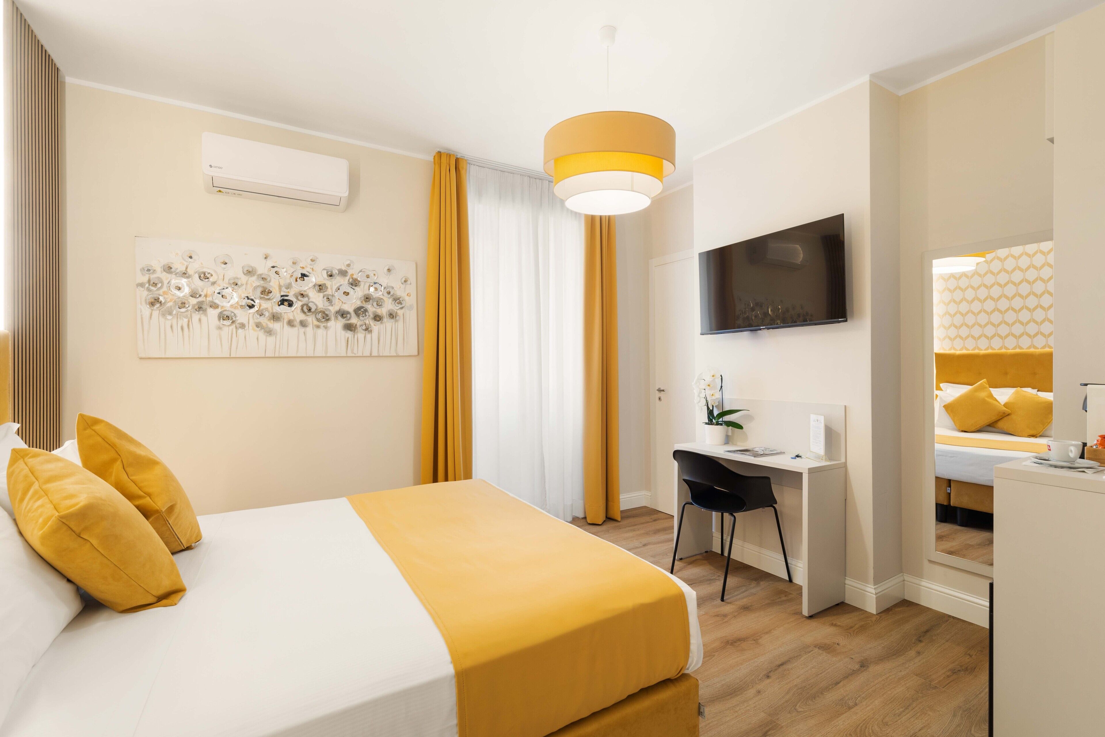 Quarto de hotel com cama, escrivaninha, cadeira, televisão e espelho.