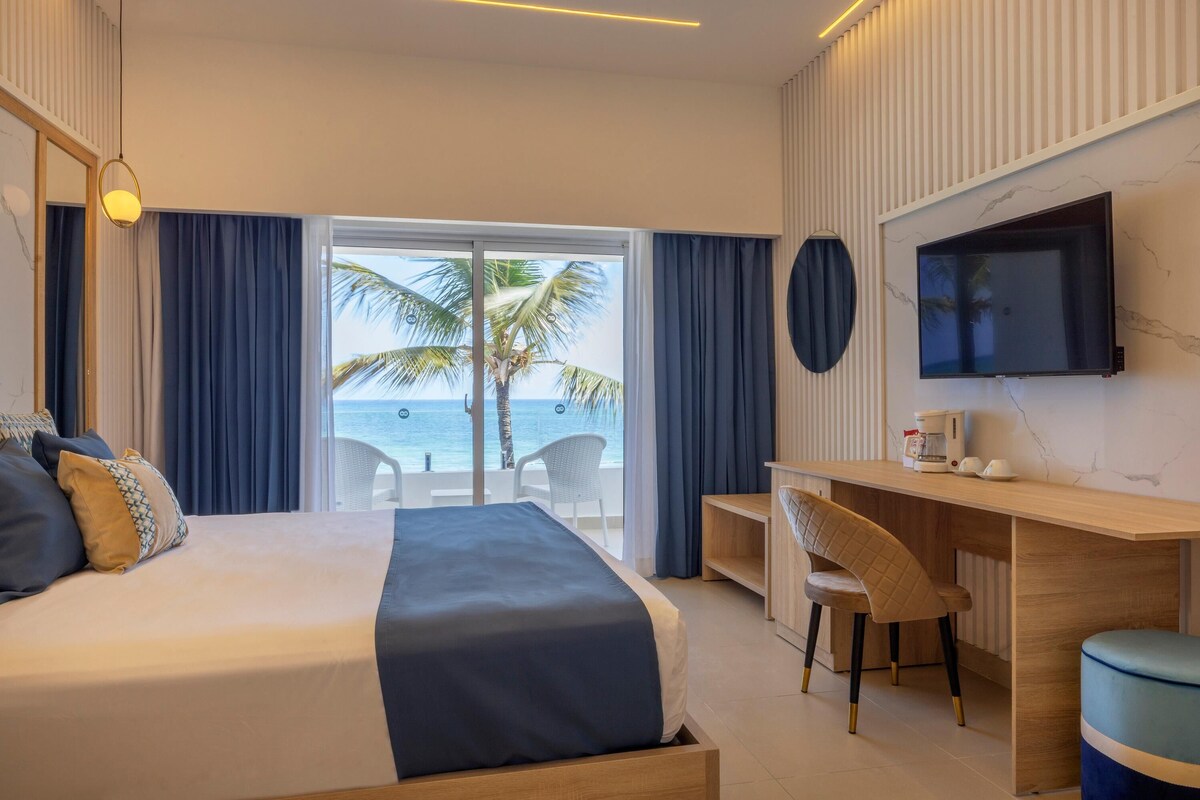 Quarto de hotel com cama, escrivaninha, cadeira, televisão e vista para o oceano.