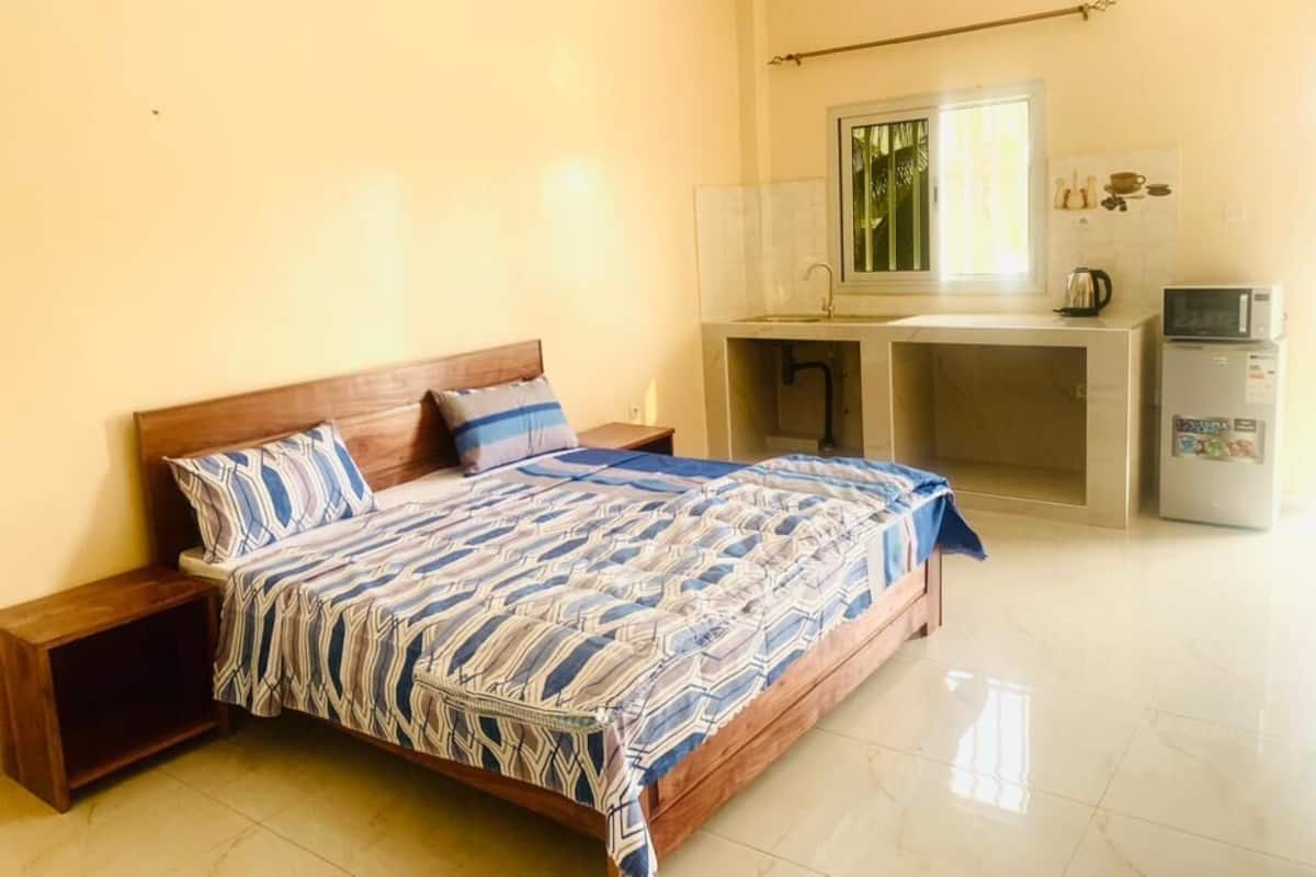 Quarto com cama, mesa de cabeceira, escrivaninha embutida com cadeira, janela e frigorífico.