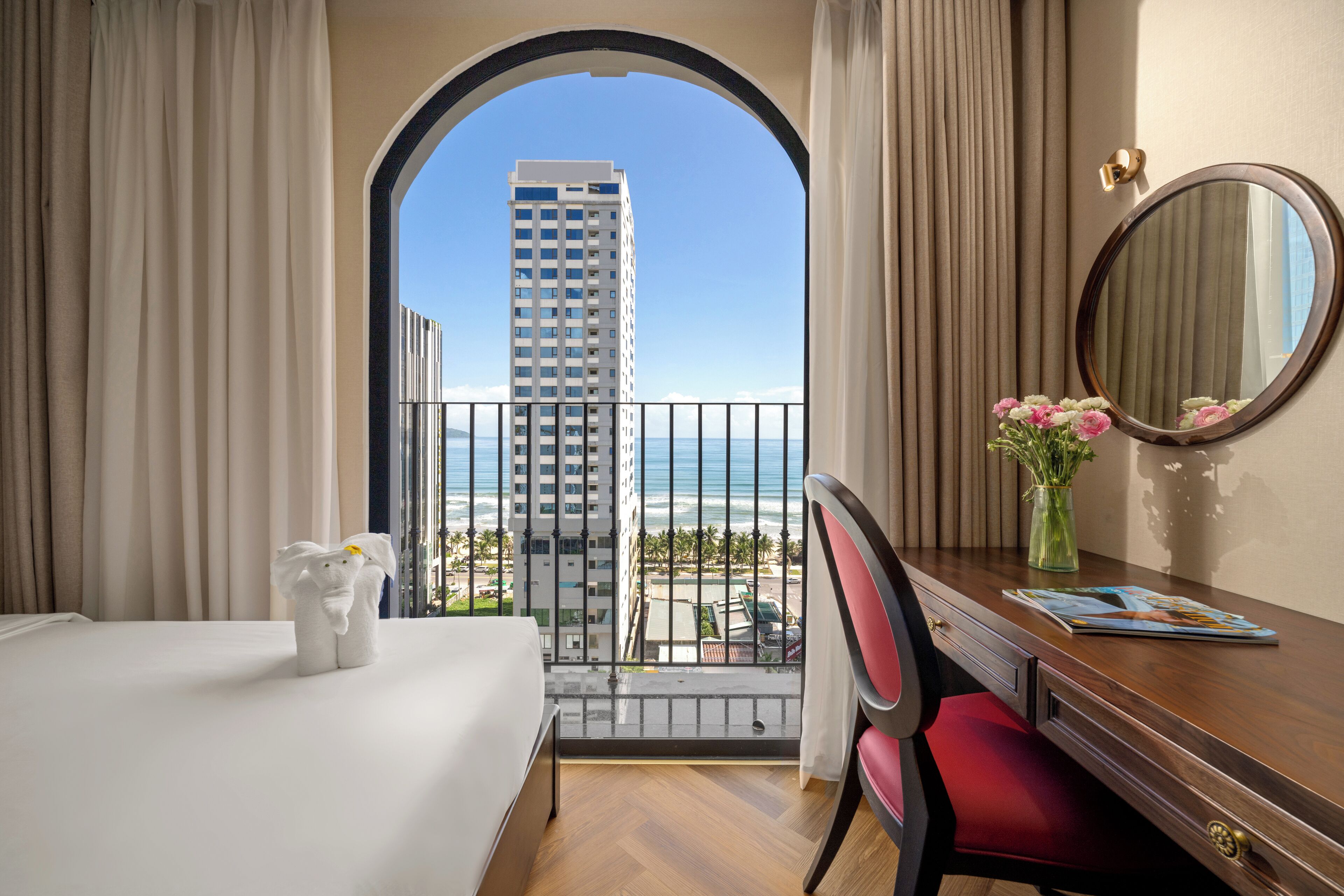 Quarto de hotel com cama, escrivaninha e cadeira, espelho e vista para um prédio alto e o oceano.