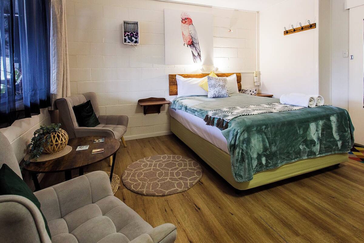 Um quarto com uma cama, uma cadeira, uma mesinha, uma prateleira na parede e uma janela com cortinas.