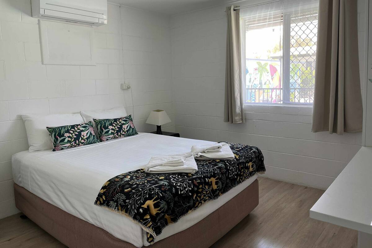 Quarto de hotel com cama, mesa de cabeceira, luminária, ar condicionado e janela com cortinas.