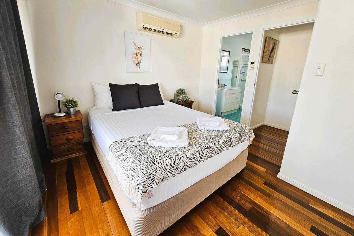 Quarto bem arrumado com cama, mesas de cabeceira, luminária e ar condicionado fixado na parede, além de um quadro emoldurado.
