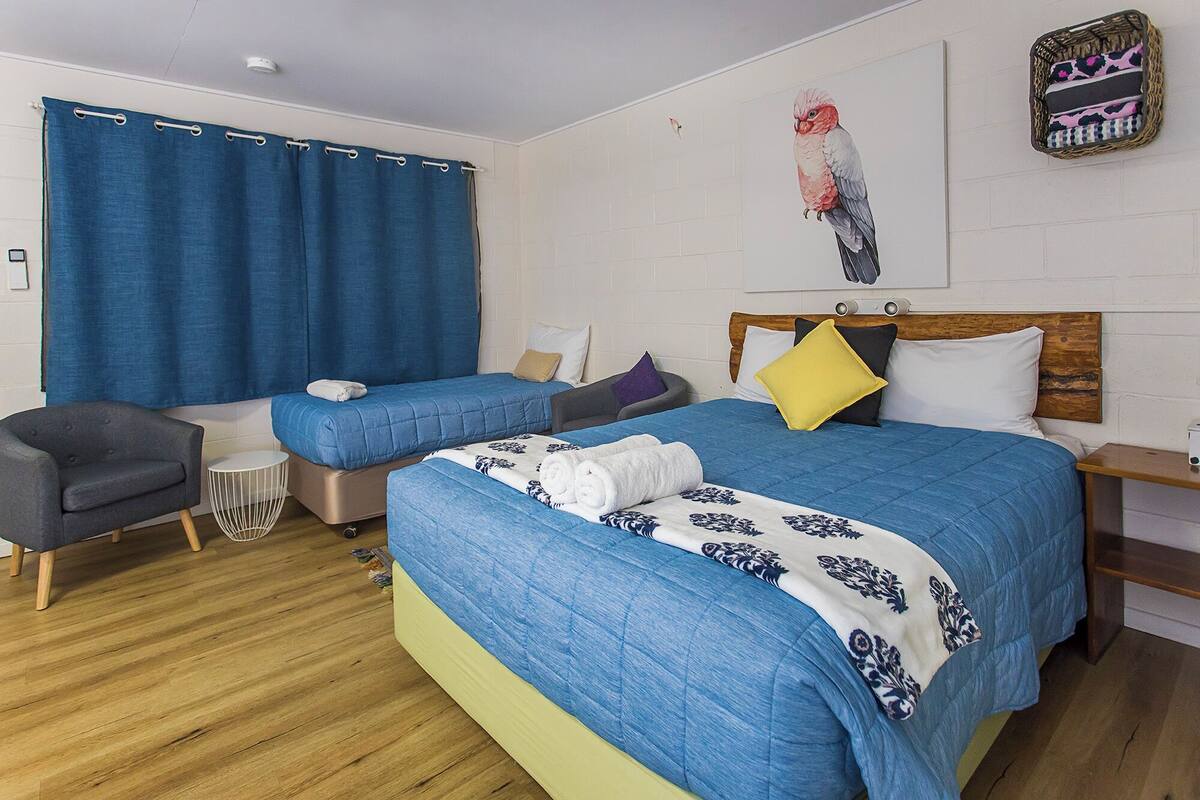 Quarto com uma cama grande, uma cama menor, uma cadeira, mesa lateral e uma obra de arte na parede.