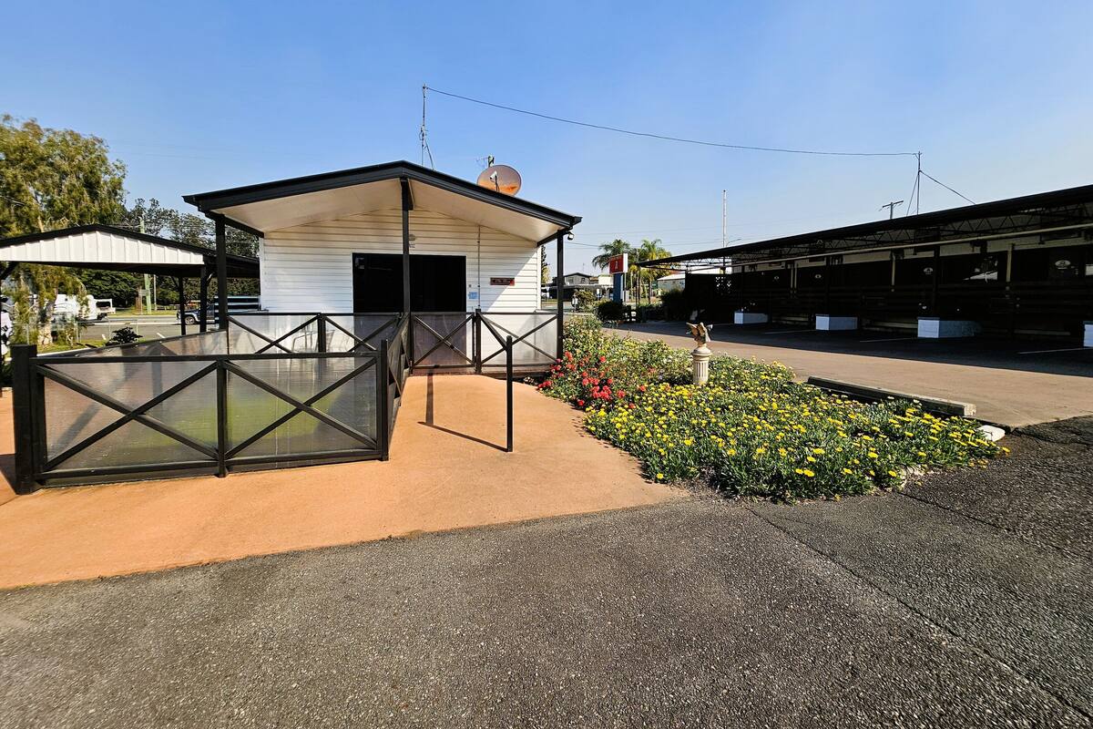 Um pequeno edifício térreo com telhado plano, entrada coberta e área cercada com flores.