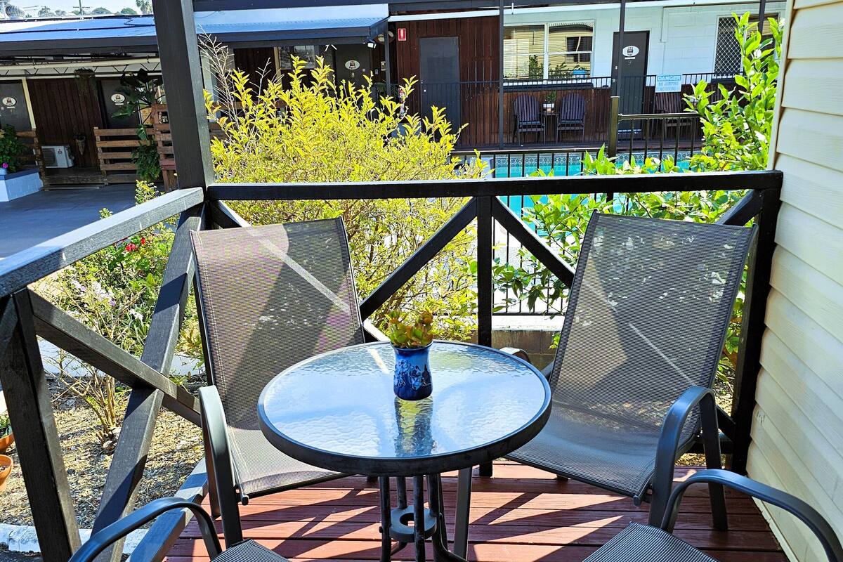 Um terraço com quatro cadeiras de rede e uma mesa de vidro, com vista para uma piscina e um edifício com varandas.