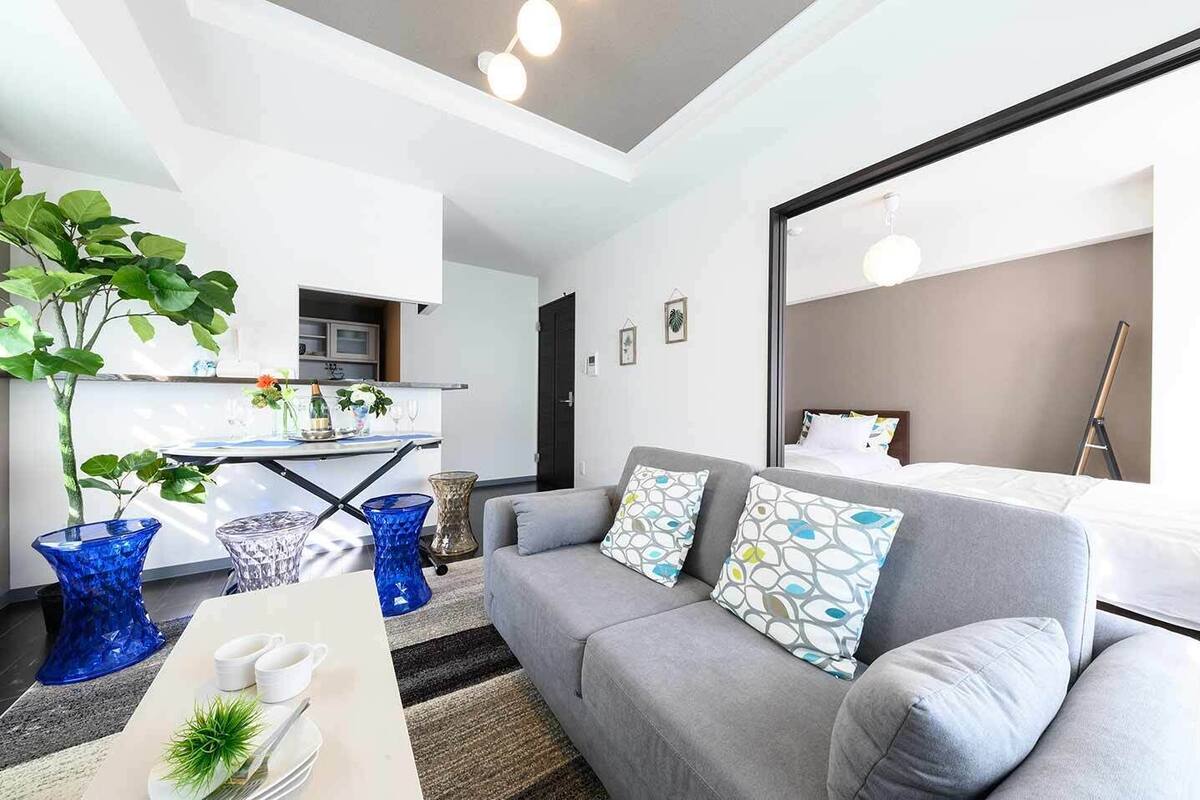Uma sala de estar moderna com um sofá cinzento, almofadas estampadas em azul e branco, uma mesa de jantar com um vaso e copos, e uma planta.