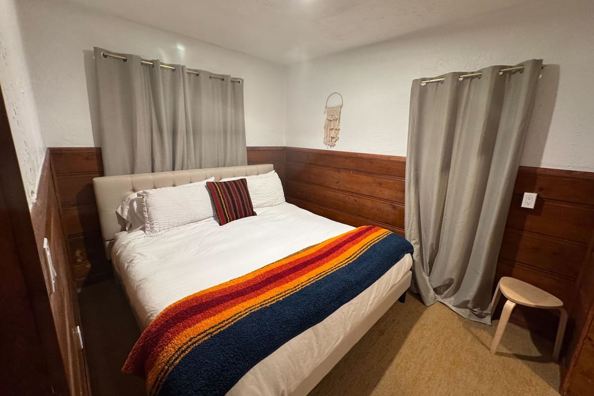 Cama bem arrumada com edredom branco e manta colorida, cabeceira de madeira, cortinas cinzas e um pequeno banquinho de madeira.