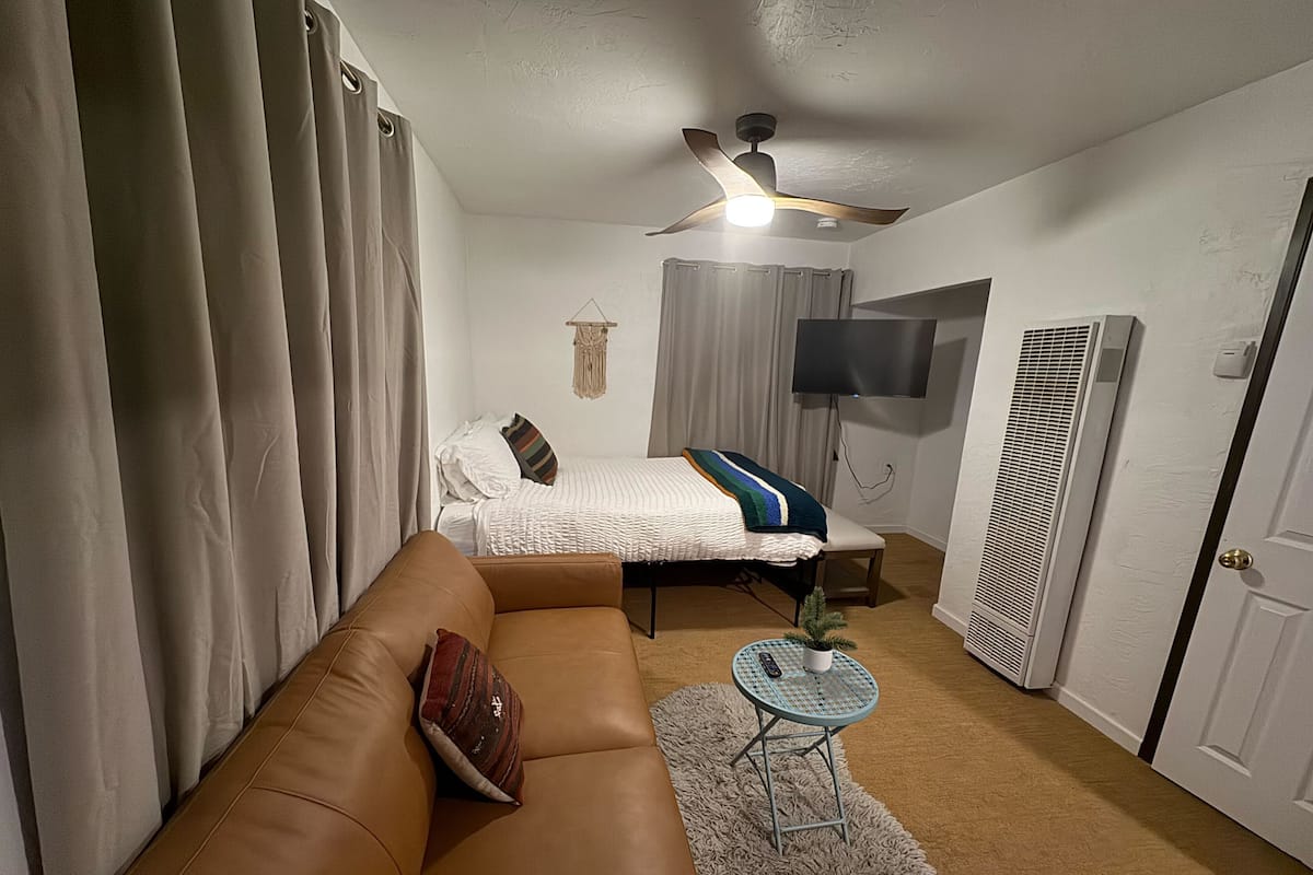 Quarto com cama, sofá, mesinha, ventilador de teto, televisão e porta.