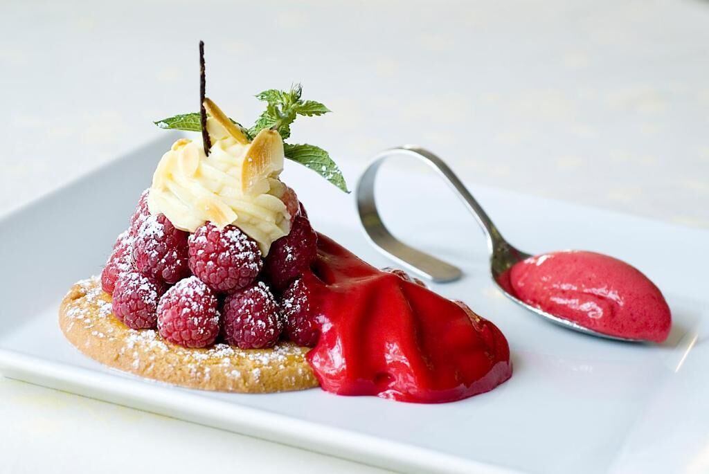 Sobremesa com framboesas, chantilly e uma cereja sobre um prato branco.