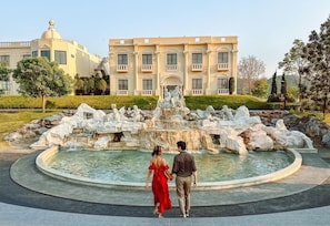 Um casal caminhando perto de uma fonte, com um edifício grande ao fundo.
