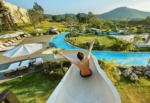 Uma pessoa escorregando em um tobogã em um resort, com piscina, cadeiras de sol e montanhas ao fundo.