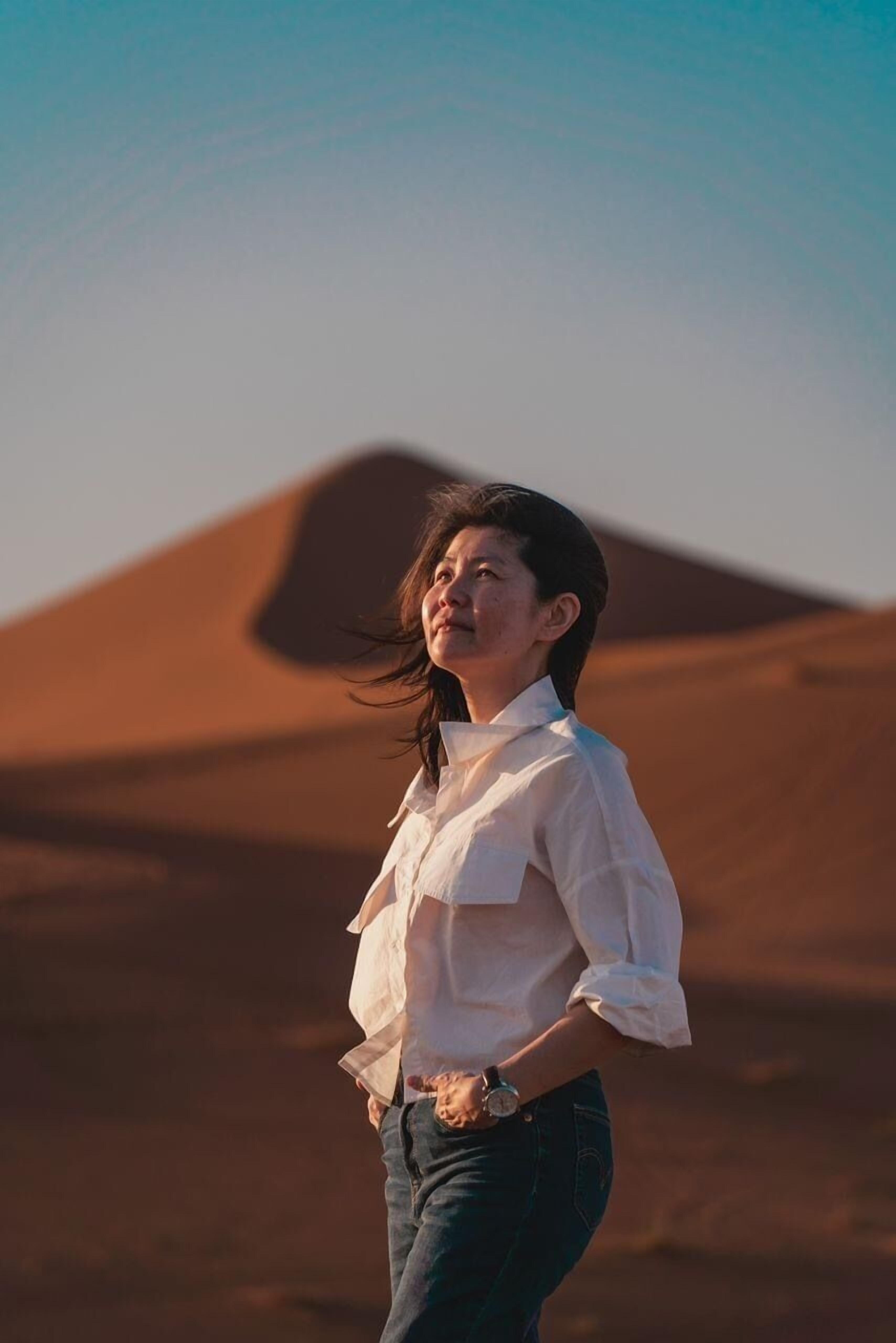Uma pessoa vestida com camisa branca e calças jeans, em paisagem desértica com dunas ao fundo.