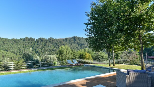 Piscina com deck de madeira e espreguiçadeiras, rodeada por cerca de metal e árvores, com uma colina arborizada ao fundo.