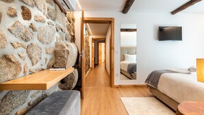 Quarto com parede de pedra, vigas de madeira, cama, televisão e corredor que leva a outro espaço.