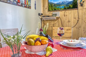 Uma mesa de jantar com uma tigela de frutas, uma taça de vinho e vista para montanhas pela janela.