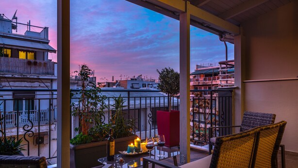 Uma varanda com cadeiras de vime, uma mesa com velas e vista para a paisagem urbana ao pôr do sol.