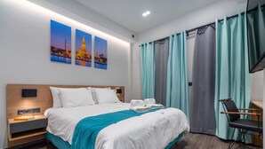Quarto de hotel moderno com uma cama grande, uma escrivaninha e uma cadeira. Há três quadros na parede, uma TV e uma janela com cortinas.