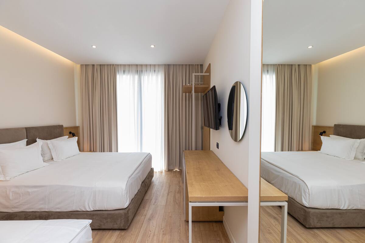 Quarto de hotel com duas camas, uma mesa de madeira e uma TV fixada na parede.