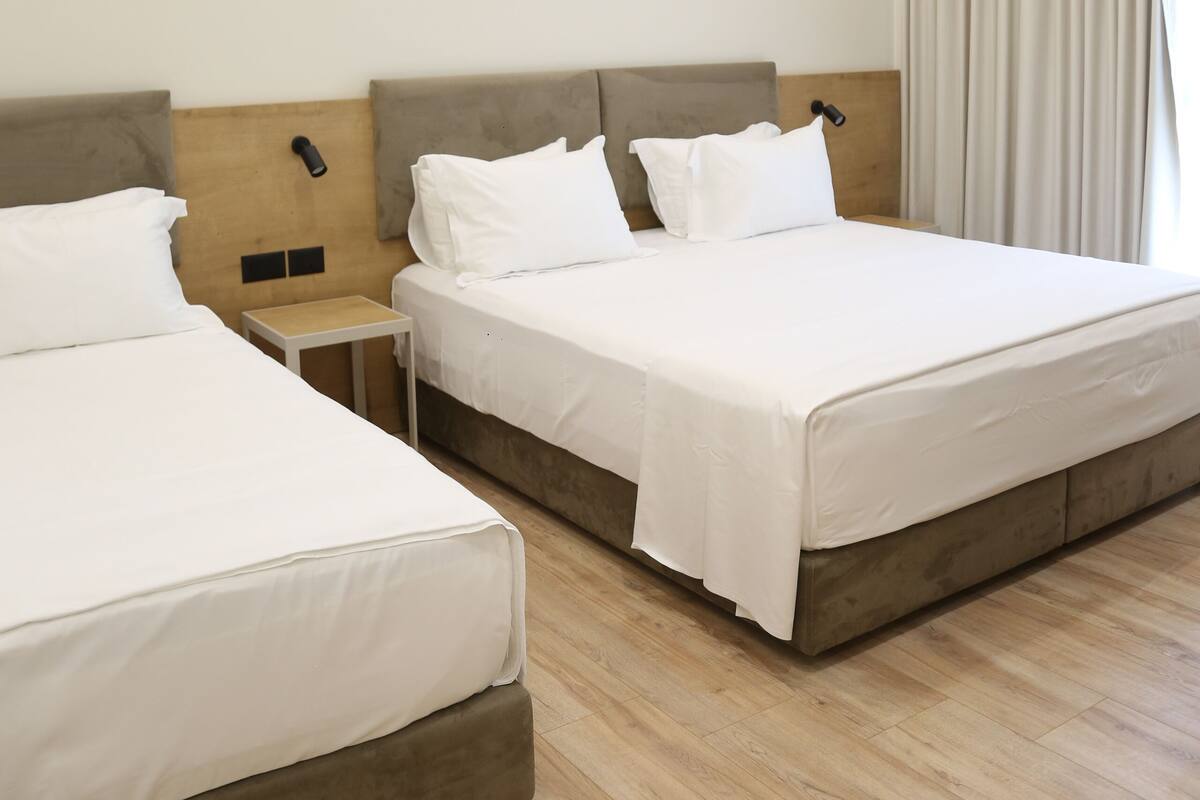 Quarto de hotel com duas camas, cabeceira de madeira, mesa de cabeceira com telefone e janela com cortinas.