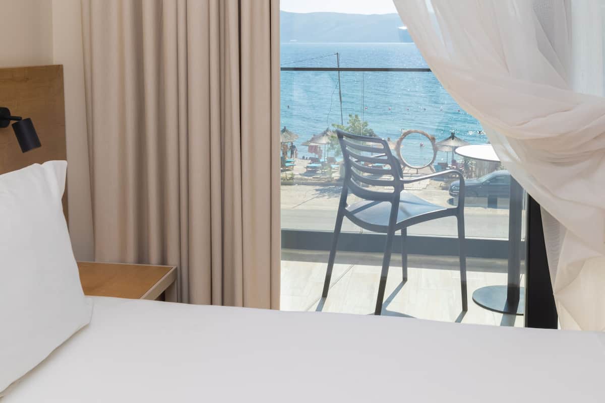 Quarto de hotel com cama, mesa de cabeceira de madeira, cadeira e varanda com vista para o mar.