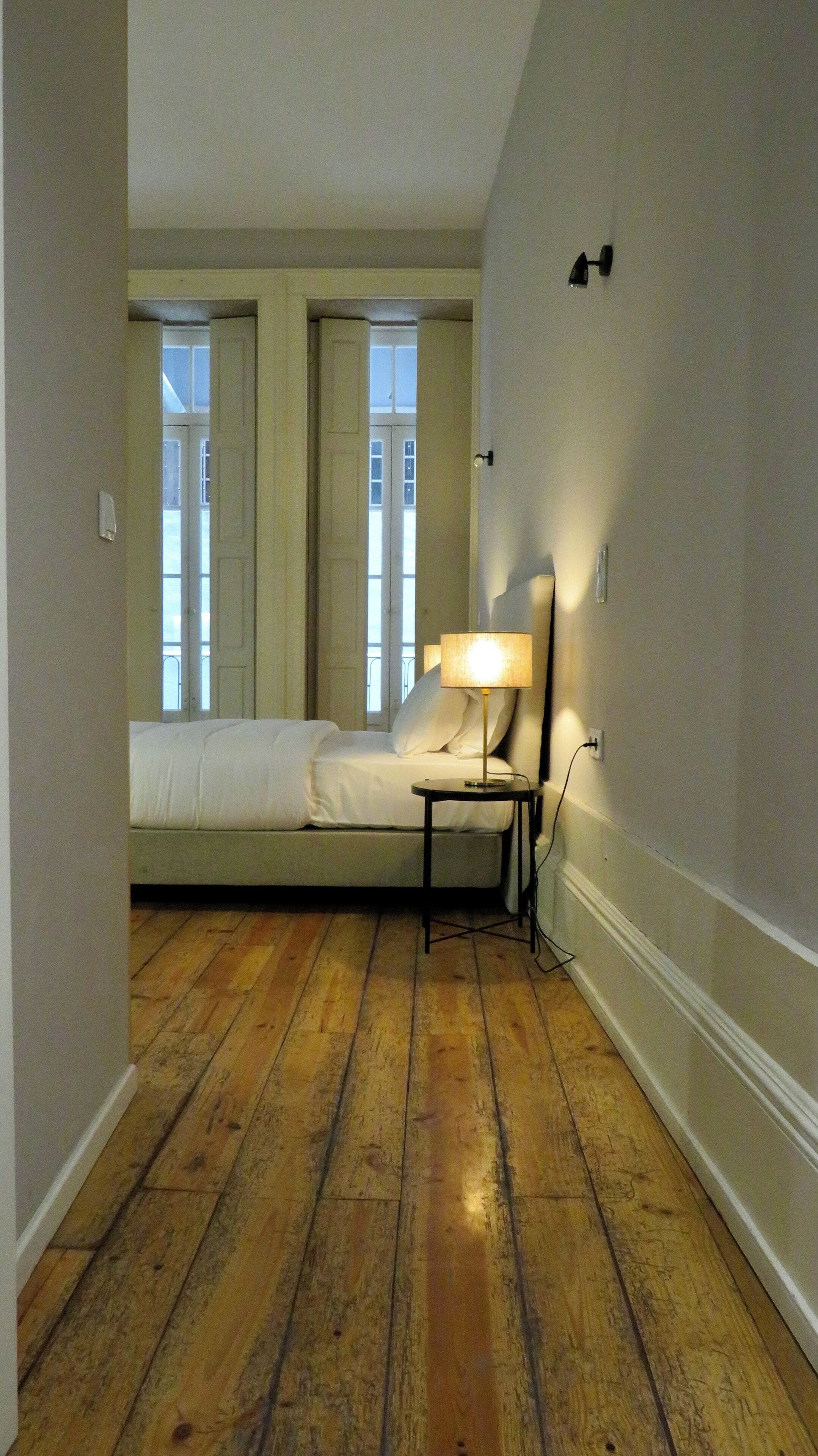 Quarto com cama, mesa de cabeceira com luminária, piso de madeira e janela com cortinas.