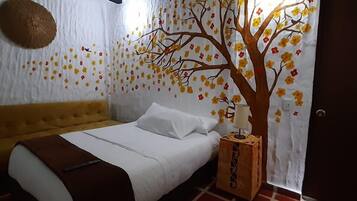 Quarto com cama, mesa de cabeceira, luminária e um mural na parede com uma árvore e folhas.
