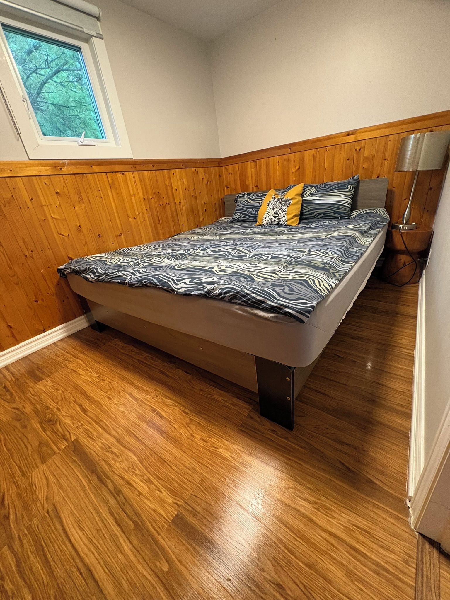 Quarto com cabeceira de madeira, cama com roupa de cama estampada, janela com vista para árvores e abajur.