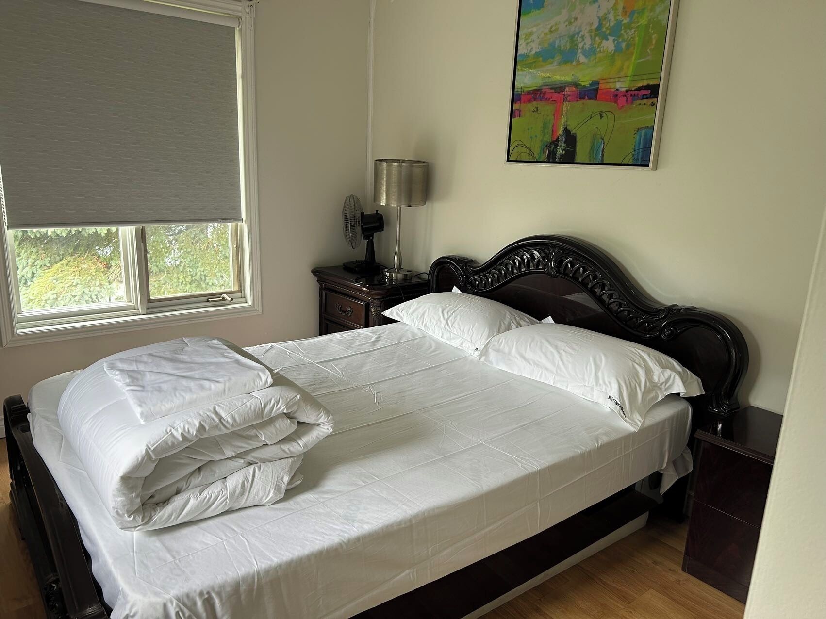 Cama bem arrumada com lençóis brancos em quarto com cabeceira de madeira, mesa de cabeceira com abajur e janela com persianas.