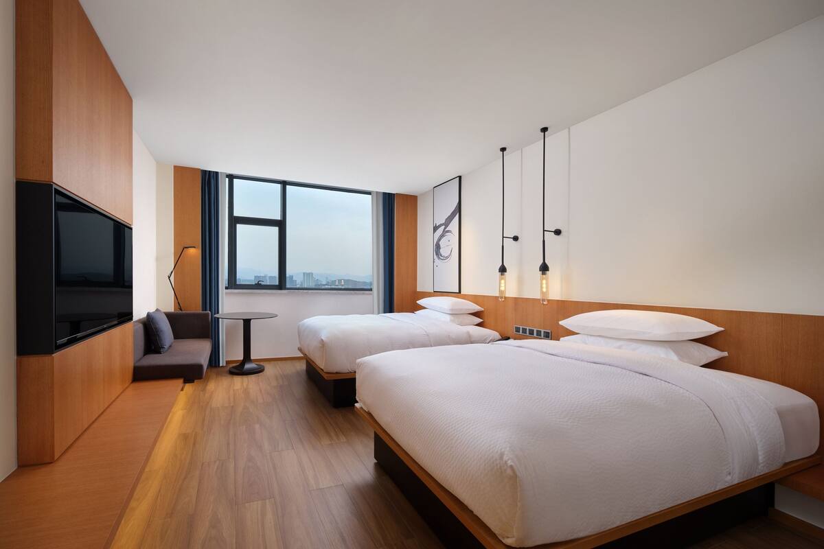 Quarto de hotel moderno com duas camas, um sofá, uma TV e uma janela grande com vista para a cidade.