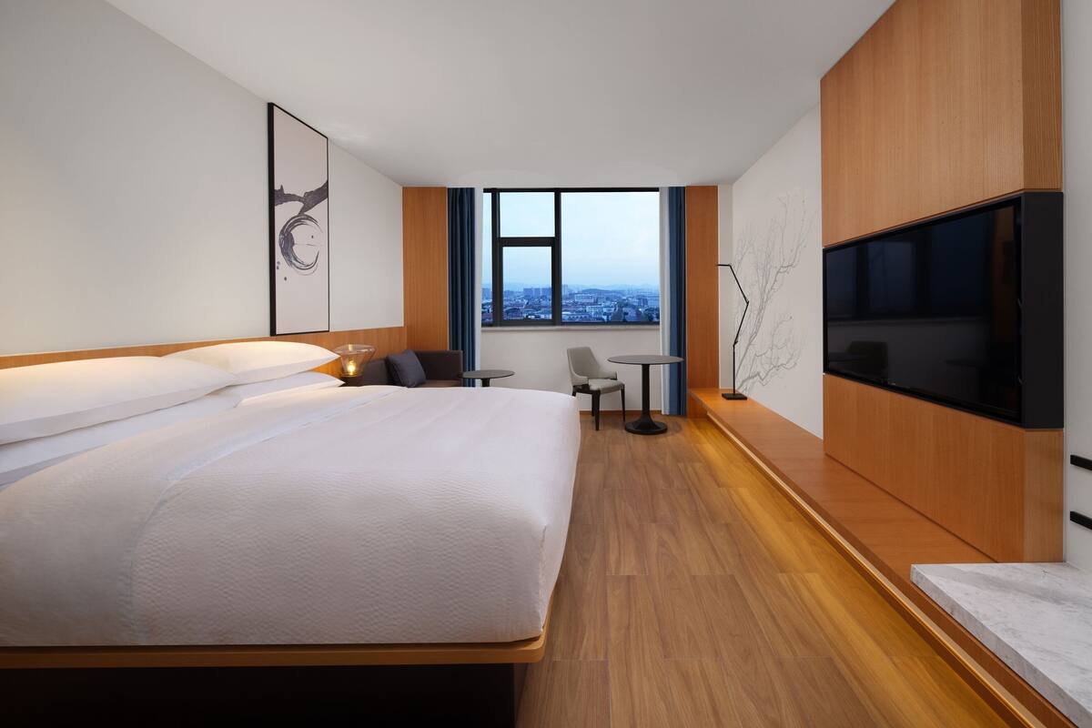 Quarto de hotel moderno com uma cama grande, televisão, escrivaninha e vista para a cidade.