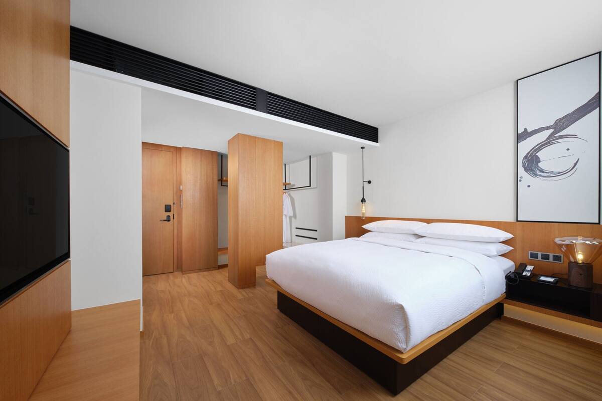 Quarto de hotel moderno com piso de madeira, uma cama grande com roupa de cama branca, mesa de cabeceira com abajur e um grande quadro abstrato na parede.