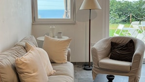 Uma sala de estar com um sofá bege, uma mesinha, um candeeiro de chão e vista para o mar através de uma janela.