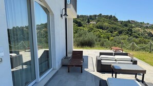 Um alpendre moderno ao ar livre com móveis de vime e uma mesa de madeira, grandes portas de vidro deslizantes e vista para uma encosta arborizada com casas.