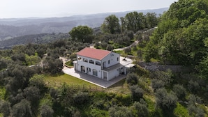 Uma casa branca de dois andares com telhado vermelho, cercada por vegetação exuberante e colinas ao fundo.