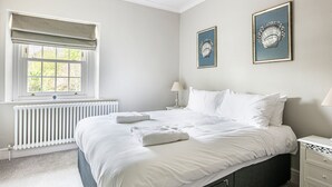 Uma cama bem arrumada com lençóis brancos, uma janela com vista para o verde e duas obras de arte emolduradas na parede.
