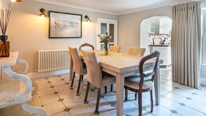 Uma sala de jantar com uma mesa grande, seis cadeiras e um vaso com flores. Há um radiador sob a mesa e um quadro na parede.