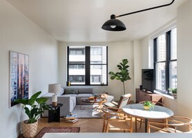 Uma sala de estar moderna com sofá, mesa de centro e área de jantar. Há uma janela grande com persianas, um vaso de planta e um quadro na parede.