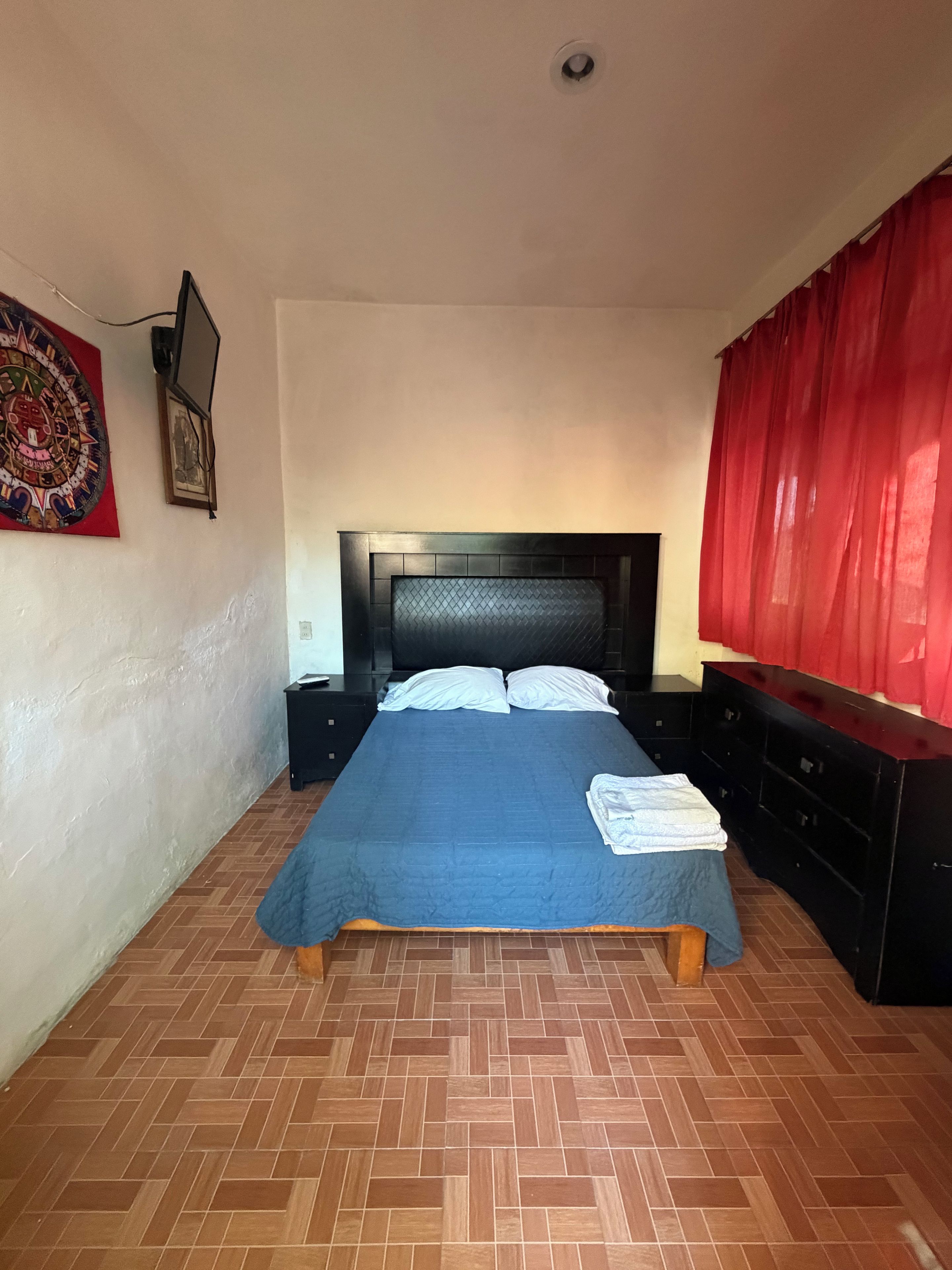 Quarto com cama, mesa de cabeceira, cômoda e cortina vermelha.